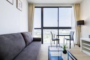 Uma sala de estar moderna com sofá, mesa de centro de vidro, mesa de jantar com cadeiras e vista através de grandes janelas.