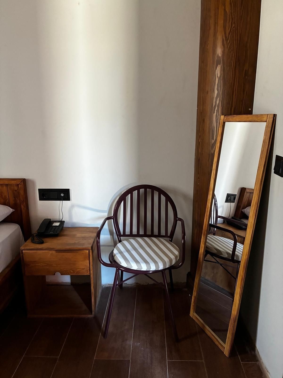 Quarto pequeno e bem arrumado com mesa de cabeceira de madeira, cadeira com almofada listrada, espelho grande e cama com cabeceira de madeira.