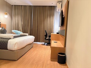 Quarto de hotel moderno com cama, escrivaninha, cadeira, televisão e lixeira.