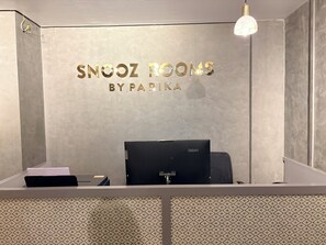 Uma mesa de recepção com um laptop, uma pasta rosa e uma cadeira preta. A parede ao fundo tem uma placa metálica com a inscrição 'SNOOZ ROOMS BY PAPIKA'.