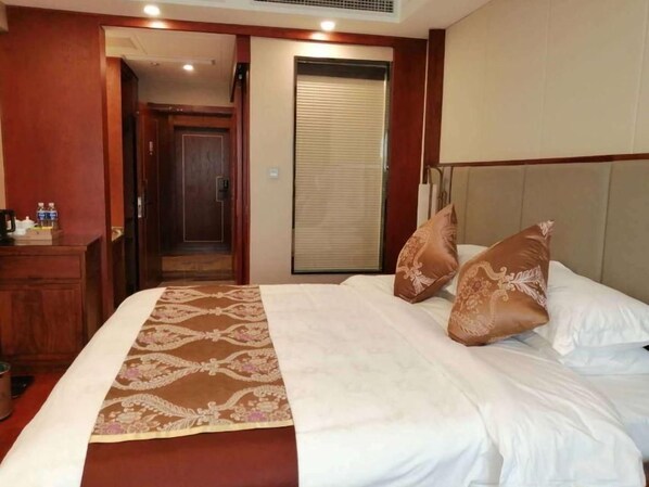 Quarto de hotel com cama, almofadas, mesa de cabeceira e porta que dá para um corredor.