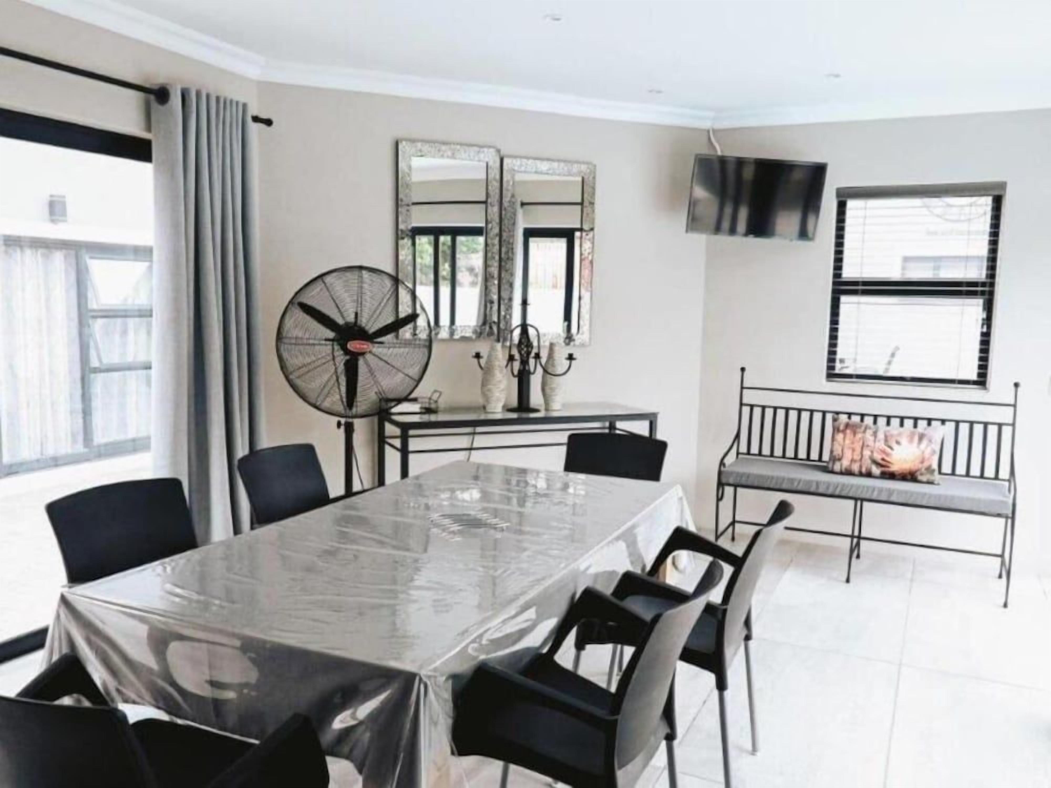 Uma área de refeições com uma mesa coberta por um lençol de proteção, cadeiras pretas, um ventilador de chão, um banco com almofadas e uma TV montada na parede.