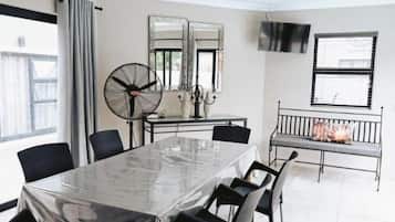 Uma área de refeições com uma mesa coberta por um lençol de proteção, cadeiras pretas, um ventilador de chão, um banco com almofadas e uma TV montada na parede.
