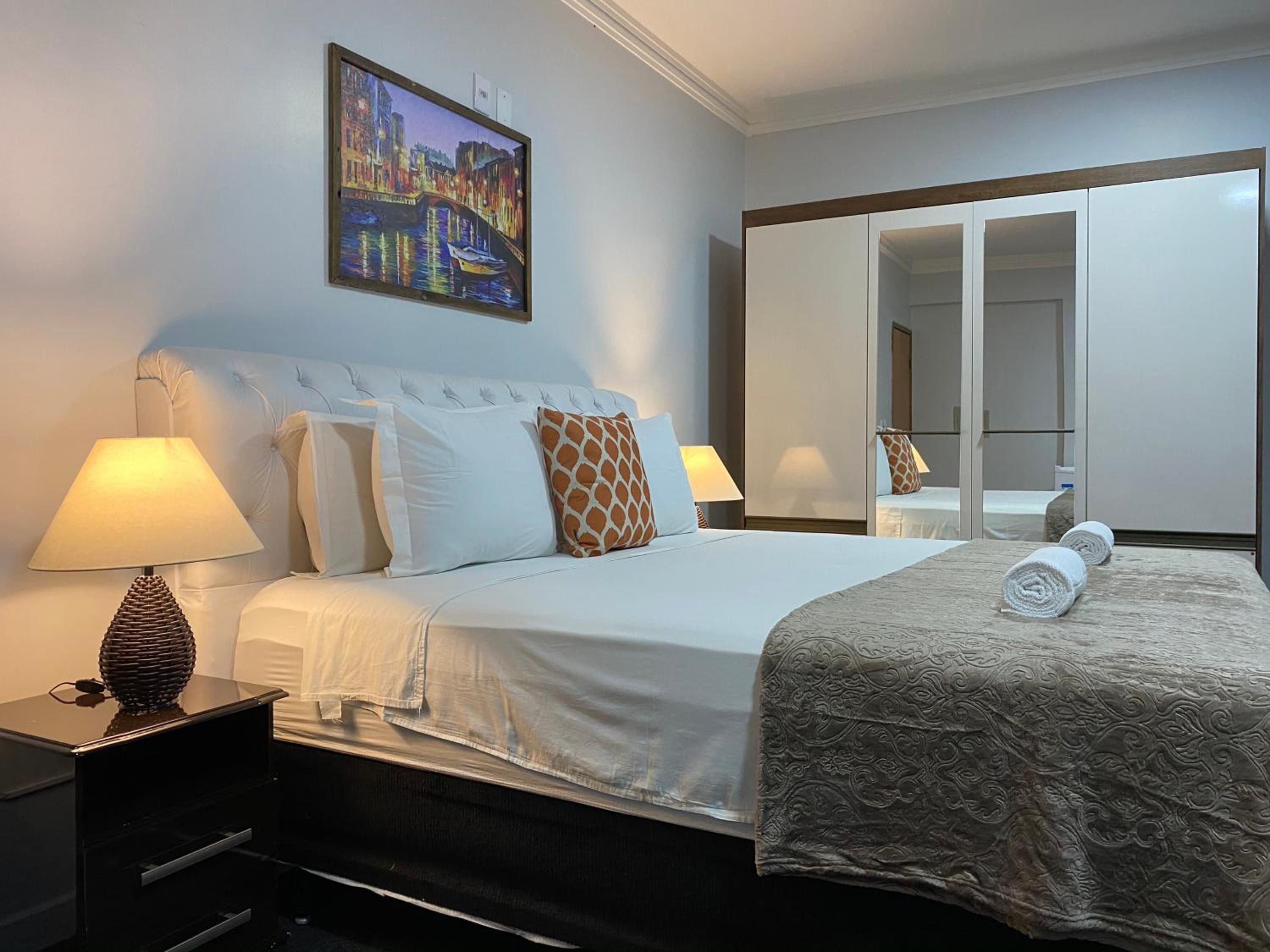 Quarto de hotel com cama, abajures, um espelho grande e um quadro na parede.