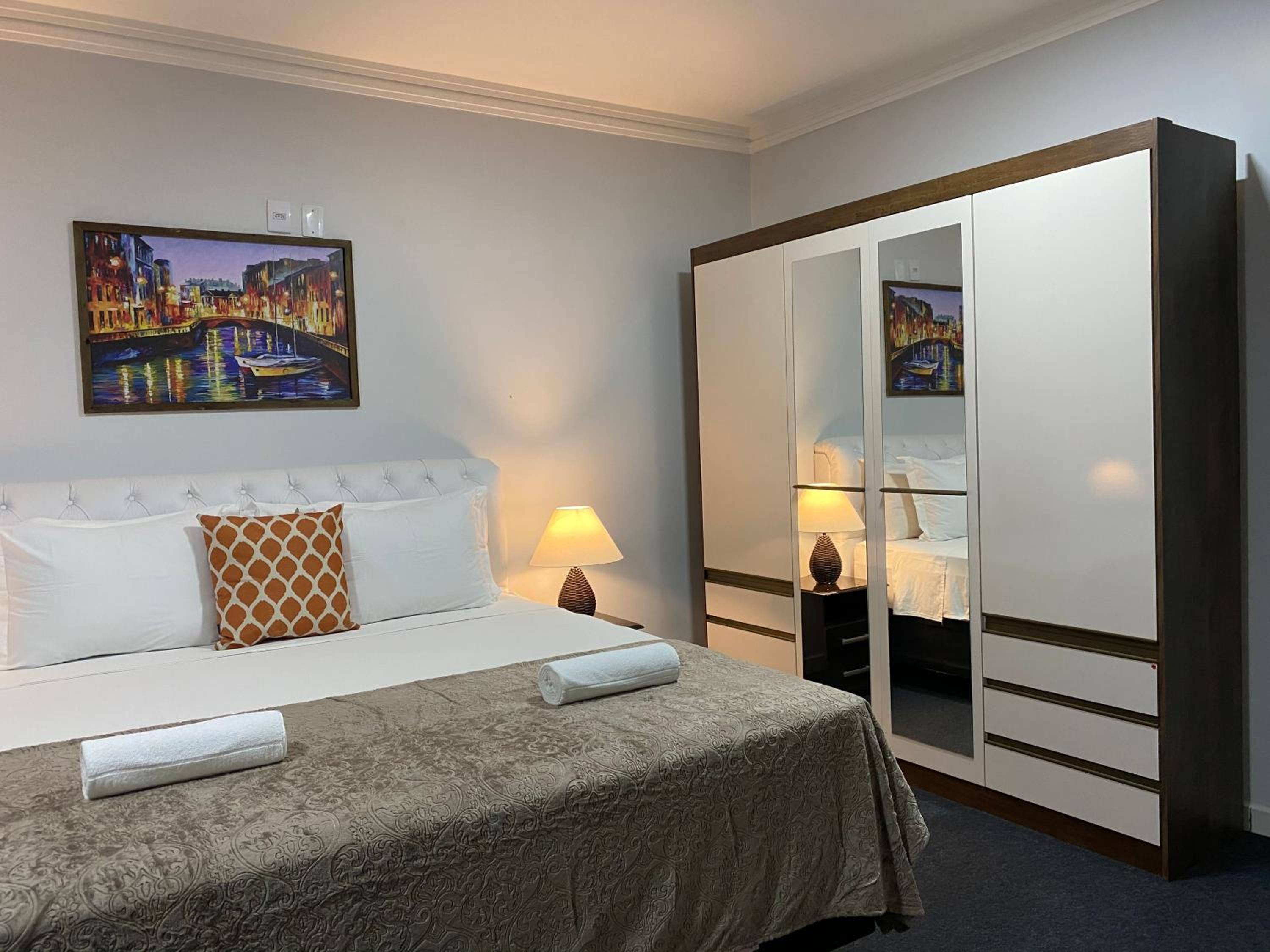 Quarto de hotel com uma cama, mesa de cabeceira, luminária, guarda-roupa e um quadro na parede.