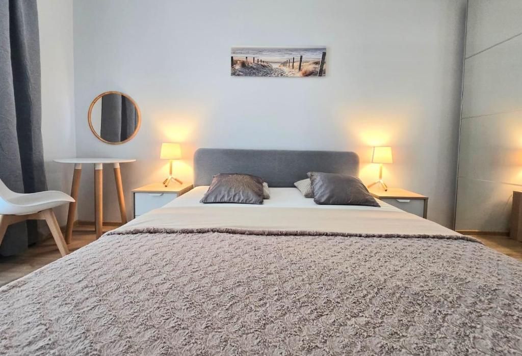 Quarto com cama, luminárias de cabeceira, espelho redondo, mesinha e um quadro na parede.