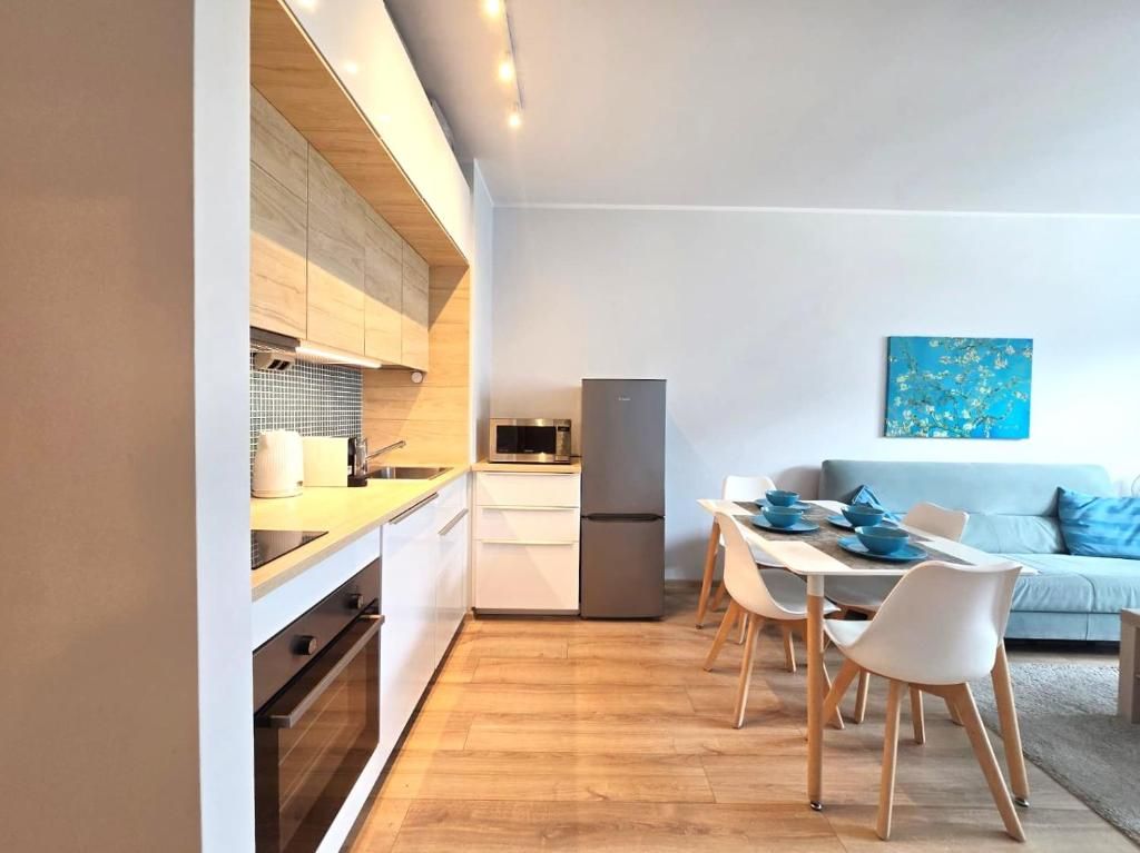 Cozinha moderna com armários brancos, forno embutido e geladeira. Há uma área de jantar com mesa e cadeiras, e um sofá ao fundo.
