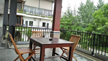 Varanda com mesa e cadeiras de madeira, guarda-corpo e vista para um edifício com varandas e vegetação.
