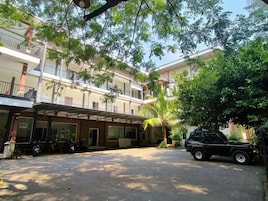 Um edifício com vários andares, entrada coberta, veículos estacionados e uma árvore ao primeiro plano.
