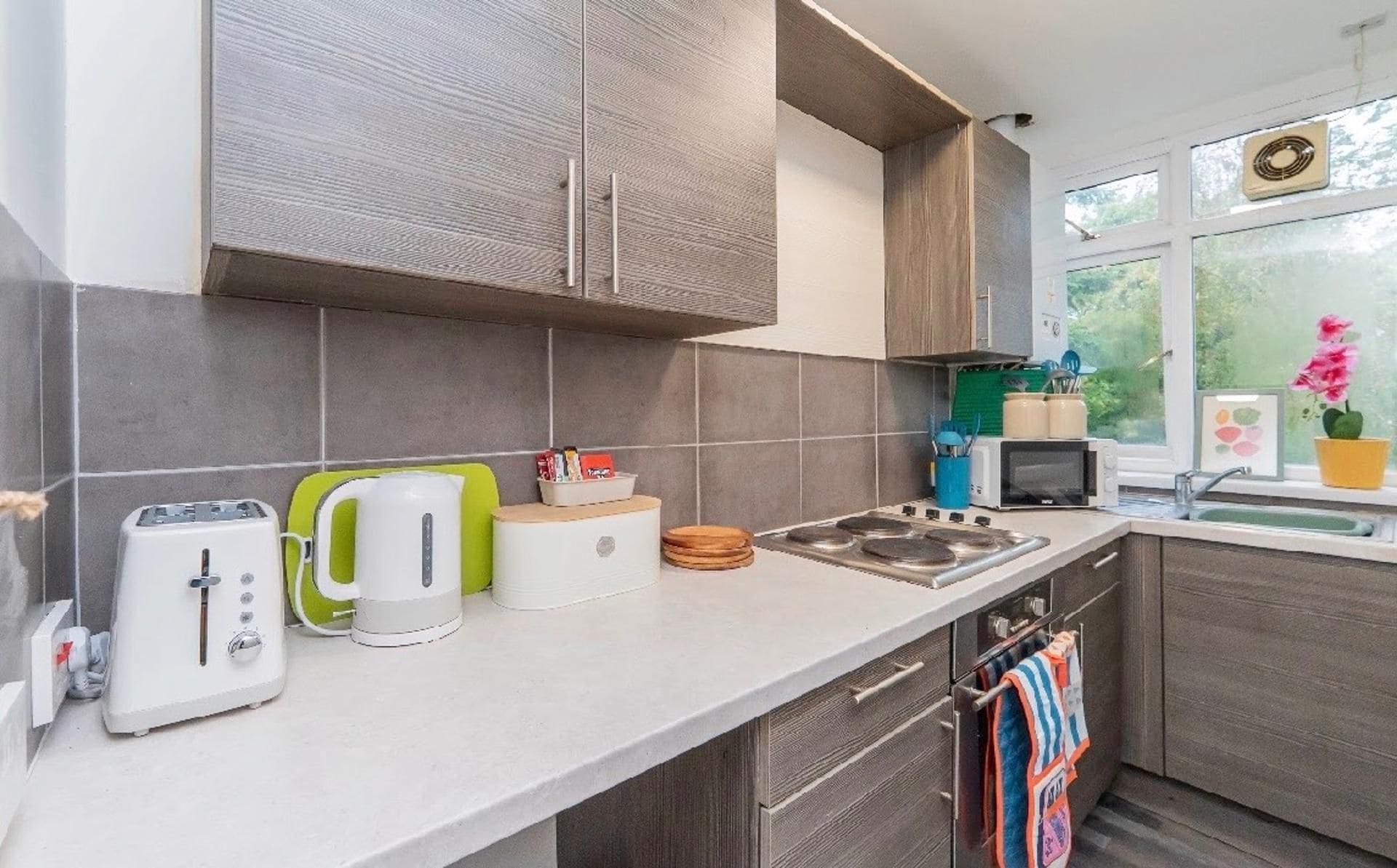 Uma cozinha moderna com armários cinza, bancada branca, torradeira, chaleira, micro-ondas e janela com vista para o verde.