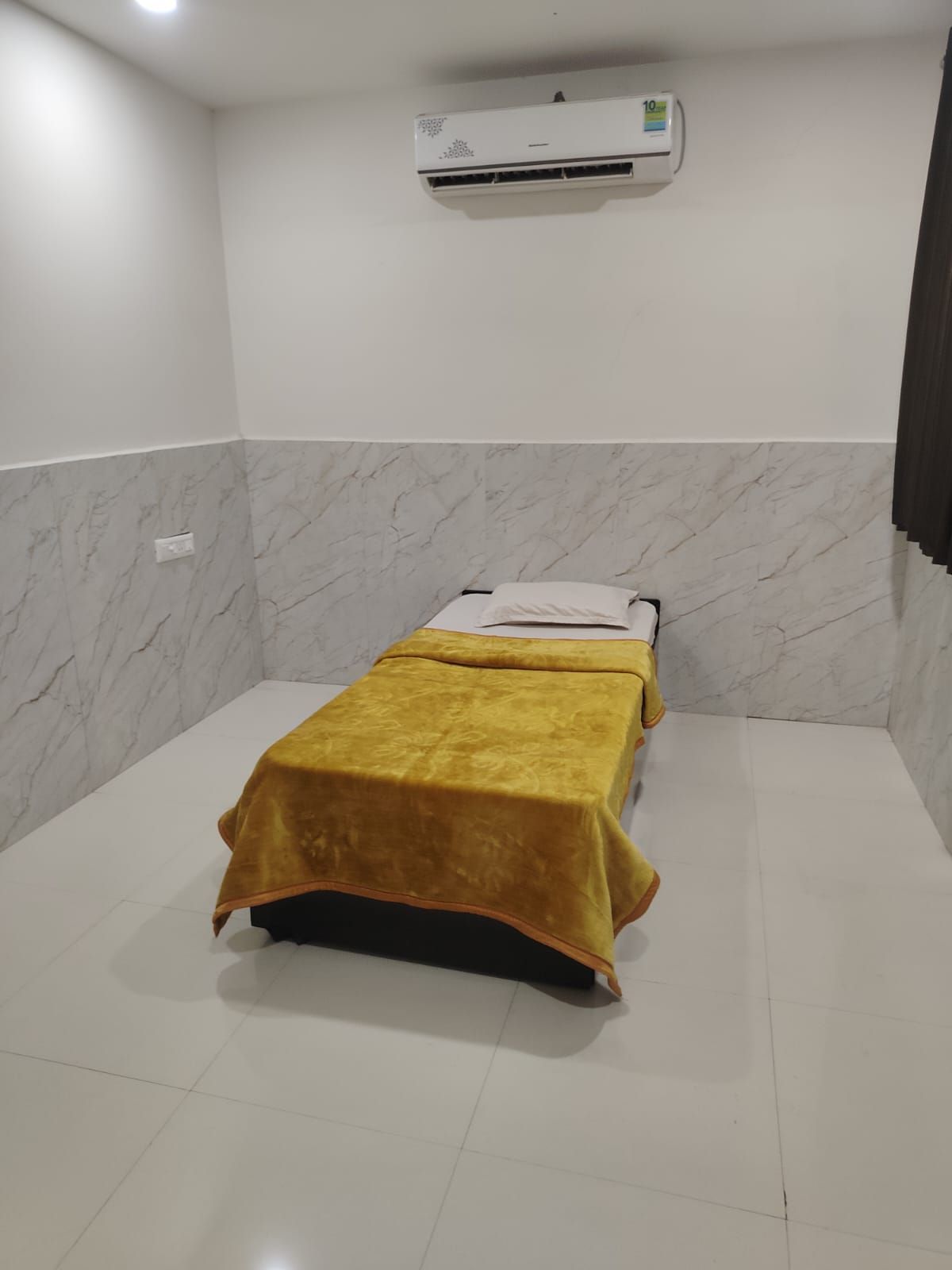 Uma cama de solteiro com colcha amarela em quarto com paredes de mármore e teto branco.