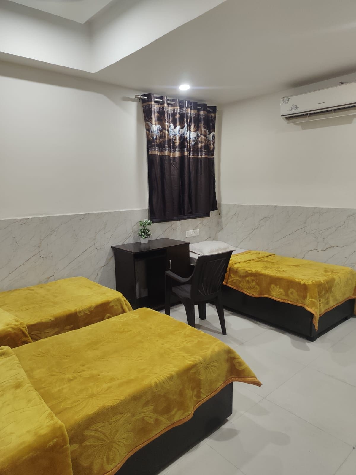 Quarto com duas camas, uma escrivaninha, uma cadeira, uma janela com cortinas e um aparelho de ar condicionado.