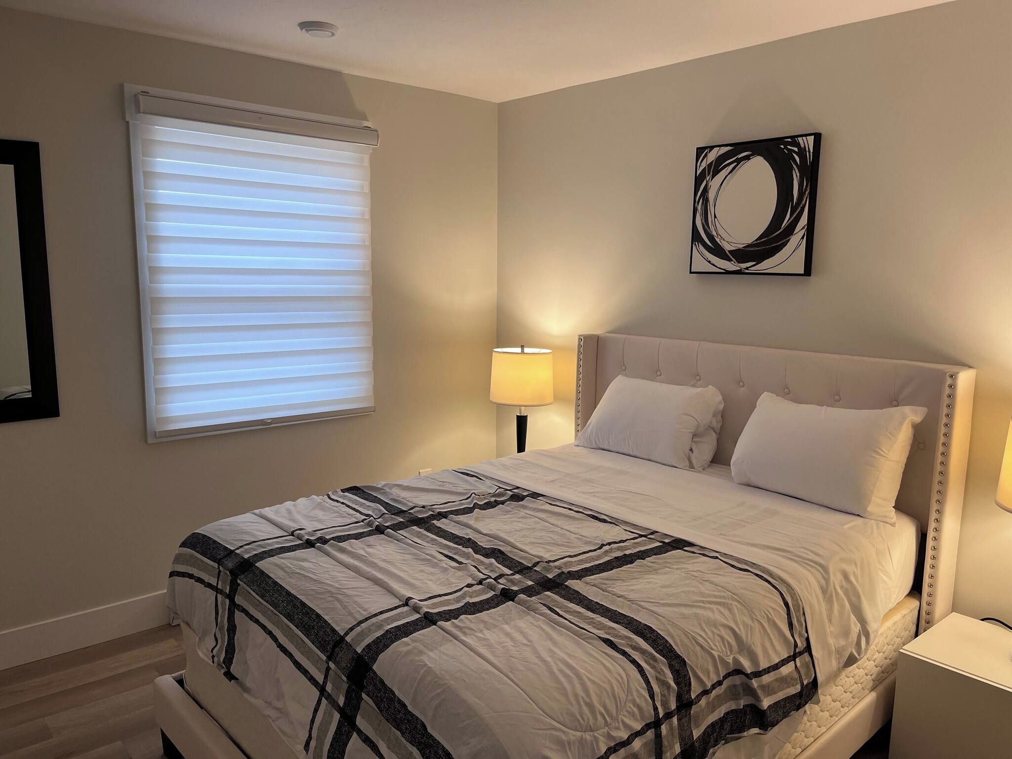 Quarto com cama, luminária de cabeceira, uma obra de arte na parede e janela com persianas.