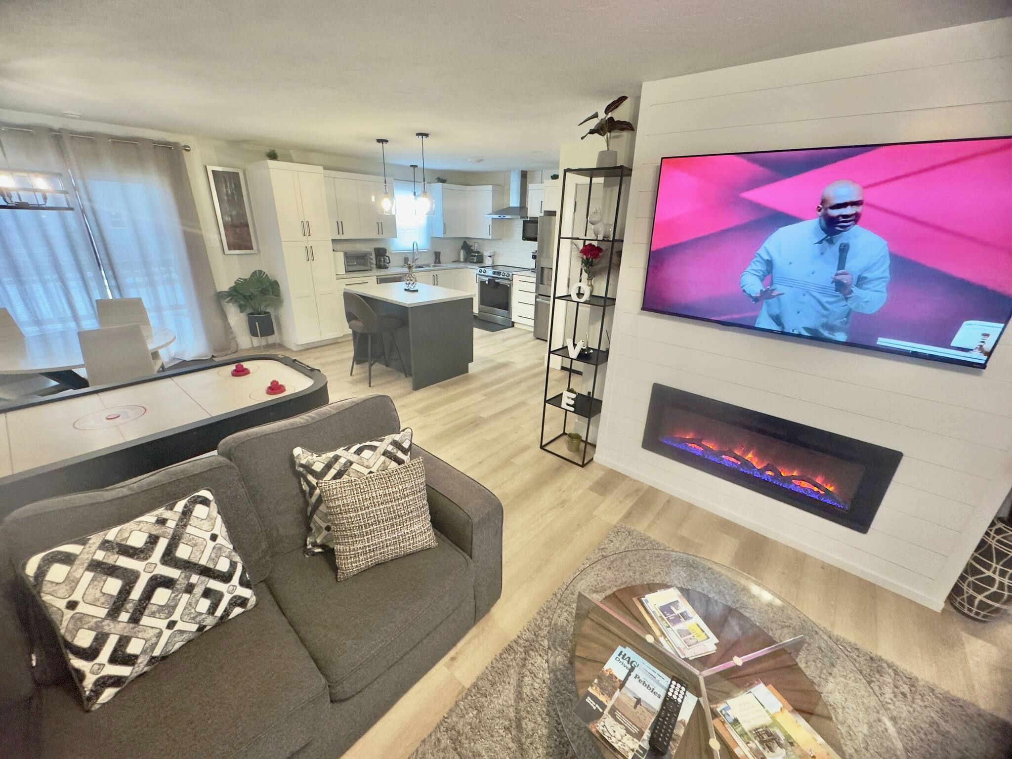 Uma sala de estar moderna com sofá cinza, televisão de tela plana na parede, lareira e cozinha ao fundo.
