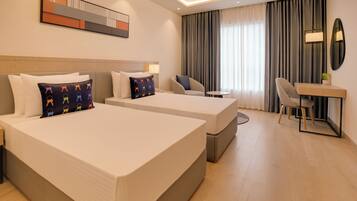 Quarto de hotel com duas camas, uma escrivaninha, uma cadeira e uma janela grande com cortinas.