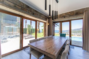 Uma sala de jantar ampla com uma grande mesa de madeira e várias cadeiras, janelas amplas com vista para uma piscina e paisagem, e uma parede de pedra.