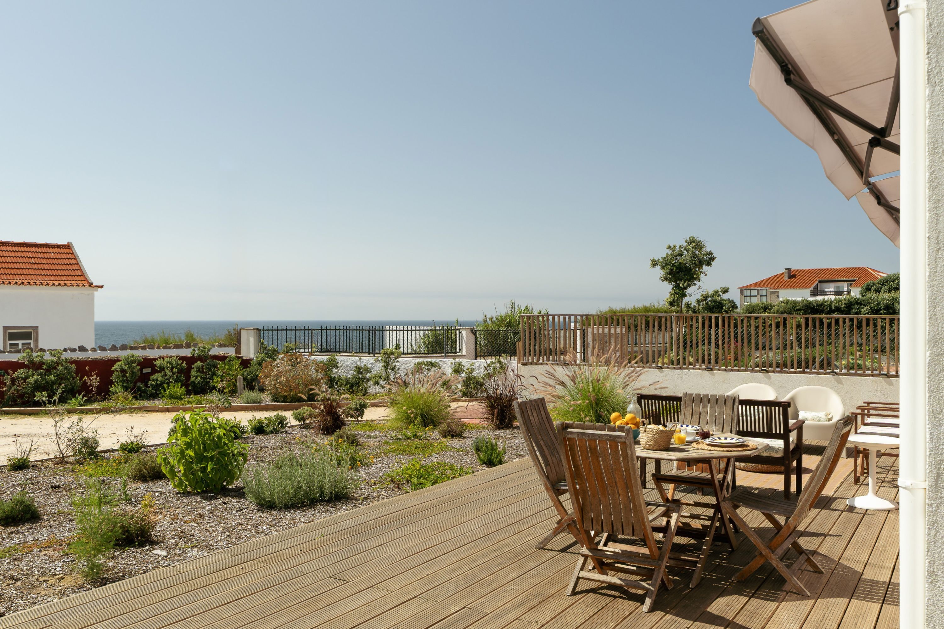 Um deck de madeira com móveis de exterior, vista para o mar e um jardim cercado.