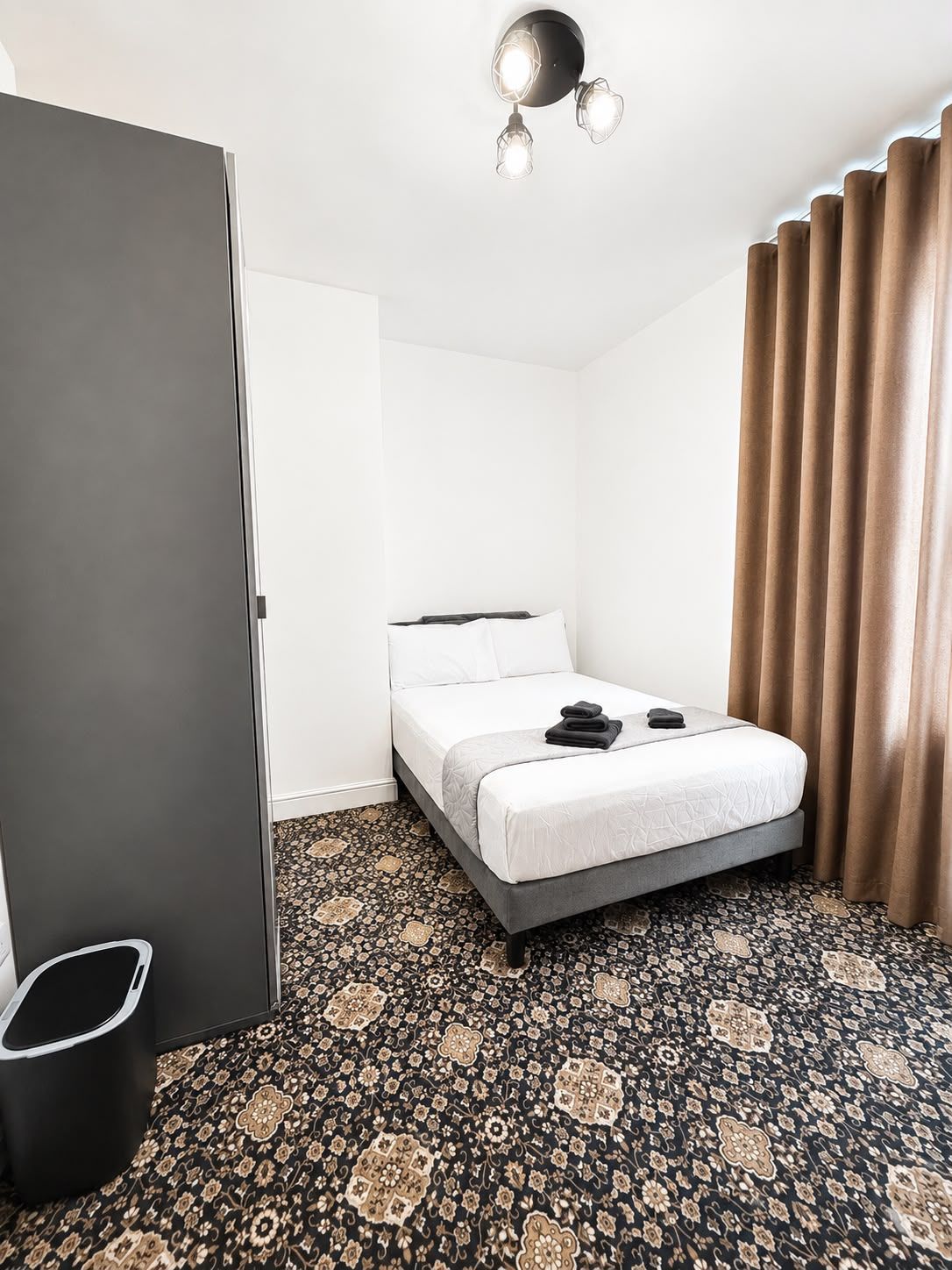 Quarto de hotel com cama, guarda-roupa, lixeira e carpete estampado.