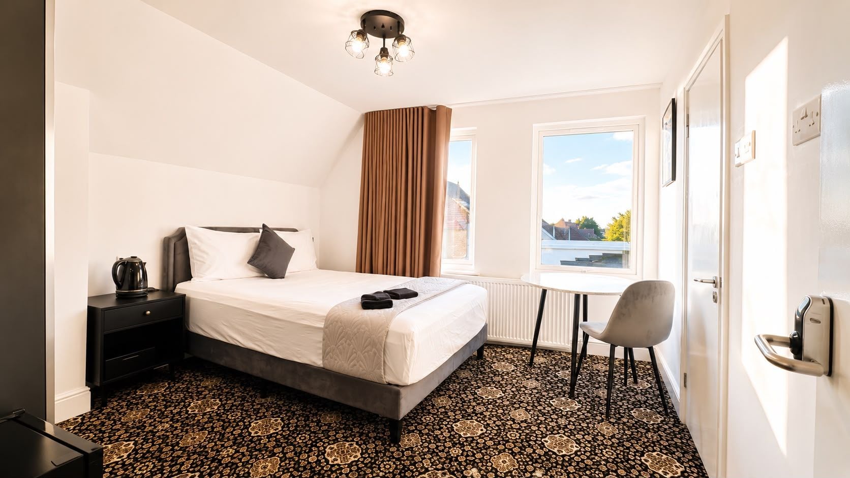 Quarto de hotel com cama, escrivaninha, cadeira, janela com vista e carpete estampado.