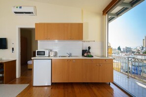 Cozinha compacta com armários de madeira, micro-ondas e pia. Há uma geladeira pequena e uma janela com vista para a cidade.