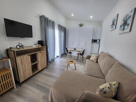 Uma sala de estar moderna com sofá, televisão fixada na parede, área de jantar com mesa e cadeiras, e uma kitchenette com micro-ondas.