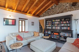 Uma sala de estar aconchegante com sofá, banquinho e uma grande estante repleta de livros e jogos de tabuleiro.