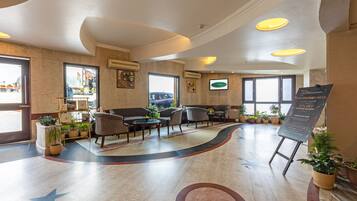 Um saguão de hotel com assentos, uma mesa de centro, plantas em vasos, uma janela grande e uma placa.