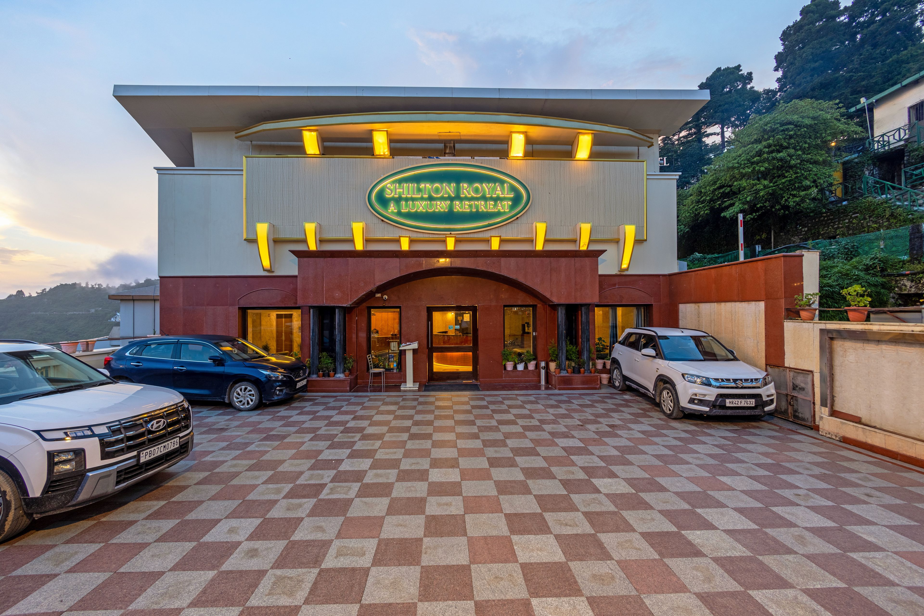 Entrada de um hotel com piso xadrez, um letreiro com a inscrição 'SHILTON ROYAL A LUXURY RETREAT' e carros estacionados na frente.