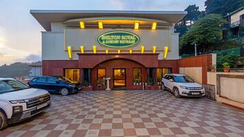 Entrada de um hotel com piso xadrez, um letreiro com a inscrição 'SHILTON ROYAL A LUXURY RETREAT' e carros estacionados na frente.