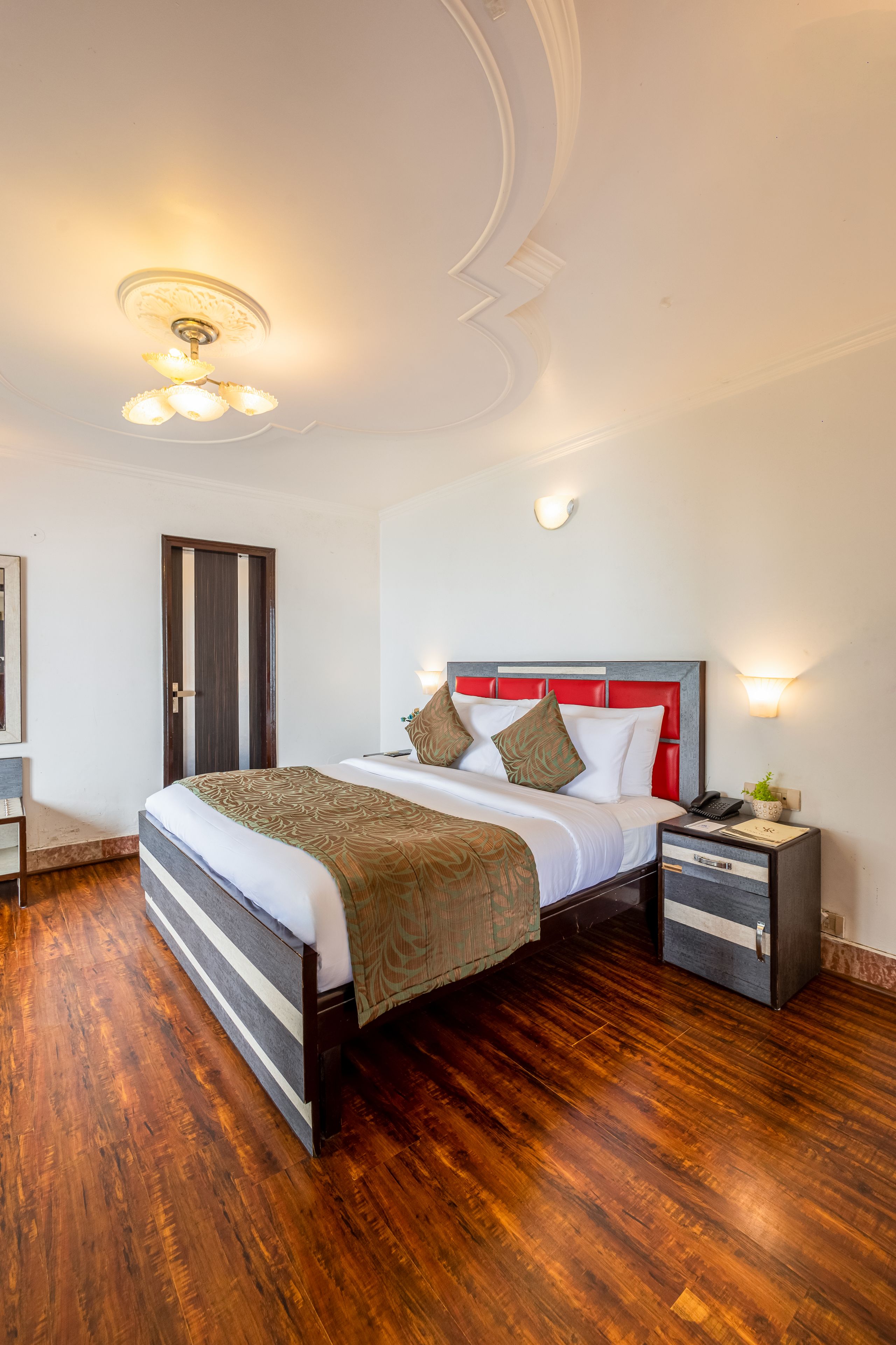 Quarto de hotel com piso de madeira, cama com cabeceira listrada, mesa de cabeceira com telefone e planta, e uma porta.