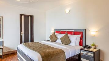 Quarto de hotel com piso de madeira, cama com cabeceira listrada, mesa de cabeceira com telefone e planta, e uma porta.