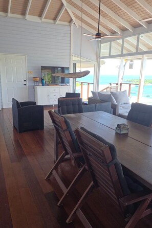 Uma sala de jantar com mesa e cadeiras de madeira, cozinha com armário branco e vista para o oceano.