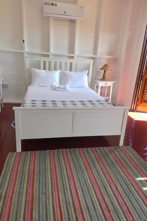 Cama branca com tapete listrado em frente, luminária de cabeceira e ar-condicionado fixado na parede.