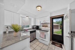 Cozinha compacta com armários brancos, micro-ondas, lava-louças e fogão. Há um vaso de planta na bancada e uma janela com vista para o exterior.