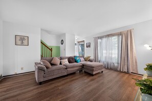 Sala de estar com sofá seccional, escada e uma grande janela com cortinas.