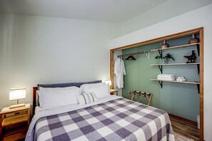 Cama bem arrumada com manta xadrez, mesa de cabeceira com abajur e prateleira na parede com toalhas e uma planta.
