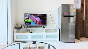 Uma sala de estar com televisão de tela plana em um suporte, unidade de entretenimento branca, mesa de centro de vidro e geladeira de aço inoxidável.