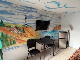 Quarto com mural, televisão de tela plana, mesa com cadeiras, geladeira e luminária.