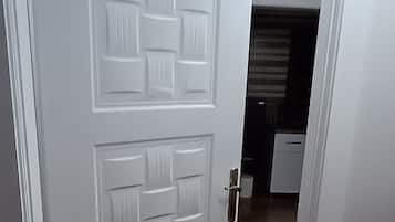 Uma porta branca com painel texturizado, dando acesso a um quarto com cozinha visível e um tapete vermelho no chão.