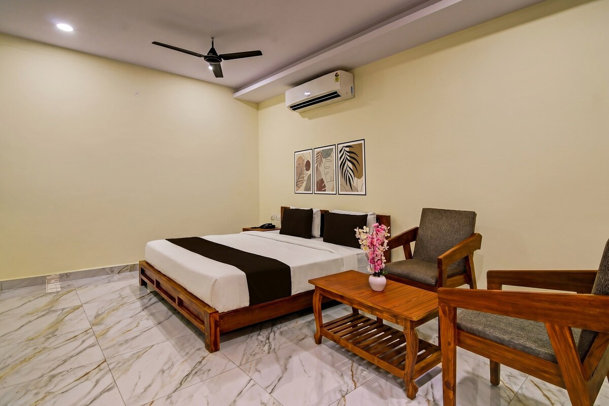 Quarto de hotel com cama, cadeira, mesa pequena, ar condicionado e ventoinha no teto.