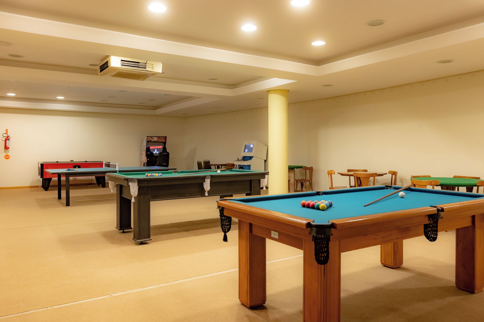 Espaço com mesa de bilhar, várias mesas, extintor de incêndio e máquina de jogos.