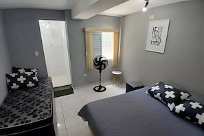 Quarto com duas camas, um ventilador, um banquinho e uma porta.