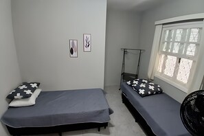 Quarto pequeno e compacto com duas camas de solteiro, cada uma com travesseiro estampado em preto e branco. Há uma janela com persianas no lado direito e um ventilador de chão no canto.