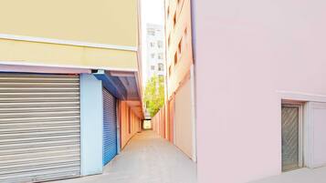 Um beco estreito entre edifícios, com persianas fechadas à esquerda e uma árvore ao centro.