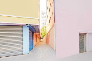 Um beco estreito entre edifícios, com persianas fechadas à esquerda e uma árvore ao centro.