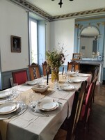 Uma sala de jantar com uma mesa comprida posta para refeição, cadeiras, um vaso com flores e uma lareira.