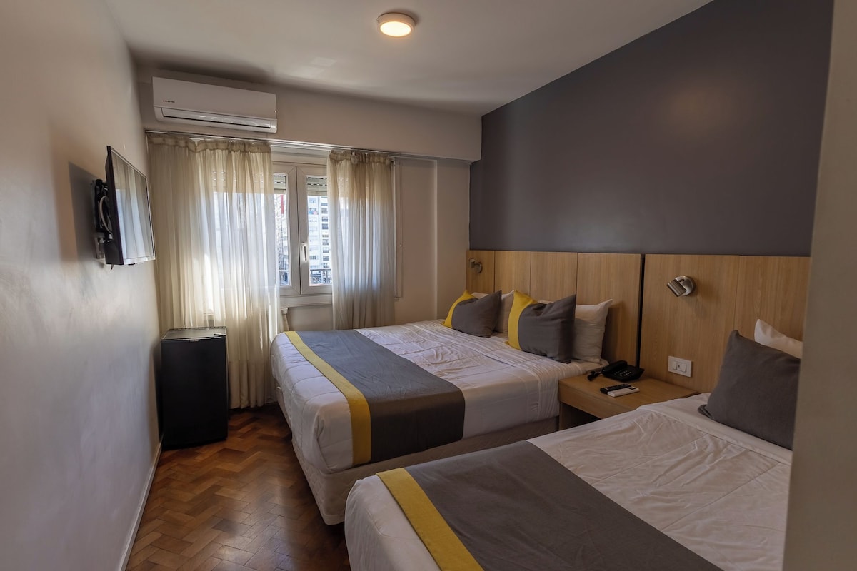 Quarto de hotel com duas camas, televisão, ar condicionado, janela com cortinas e piso de madeira.