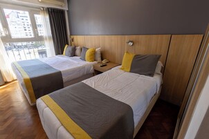 Quarto de hotel com duas camas, cabeceira de madeira, mesa de cabeceira com telefone e janela com vista para edifícios.