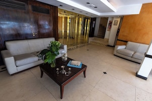 Um hall com dois sofás, uma mesa de centro com objetos decorativos e uma escada que dá acesso a um elevador.