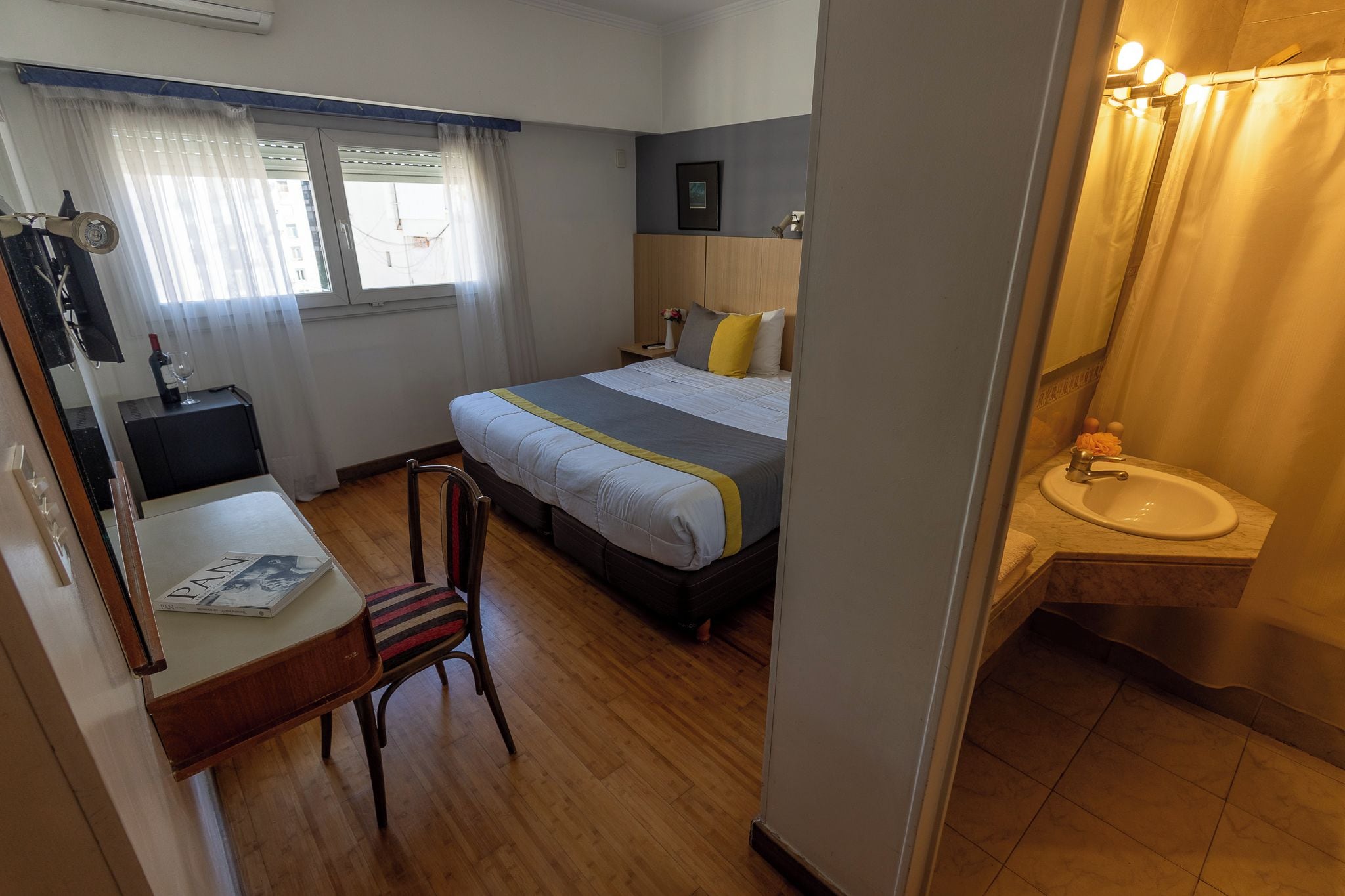 Quarto de hotel com cama, escrivaninha com cadeira, mesinha com revista, janela com cortinas e banheiro com pia e espelho.